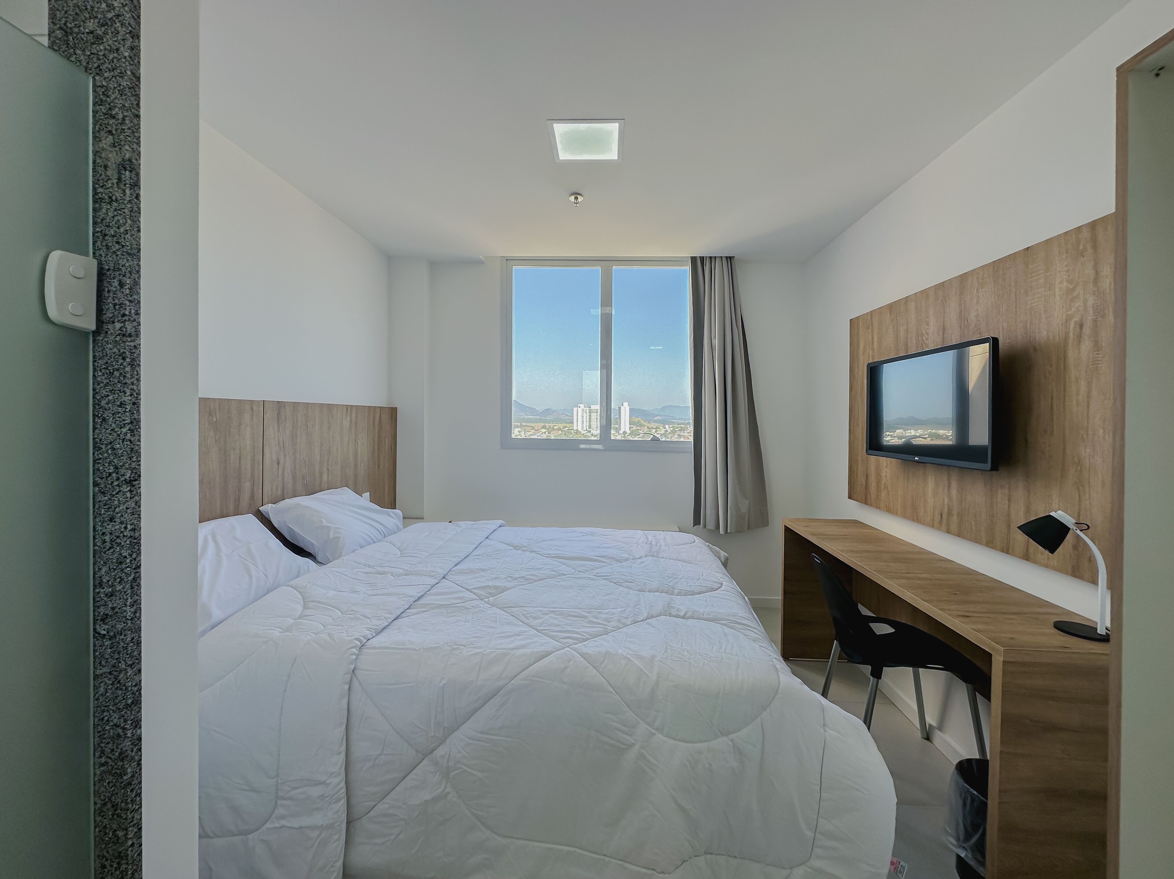 Quarto de hotel com cama, escrivaninha, televisão e janela com vista para a cidade.