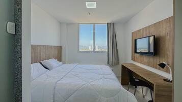 Quarto de hotel com cama, escrivaninha, televisão e janela com vista para a cidade.