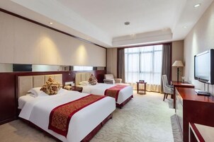 Quarto de hotel com duas camas, uma escrivaninha, televisão e uma janela grande com cortinas.