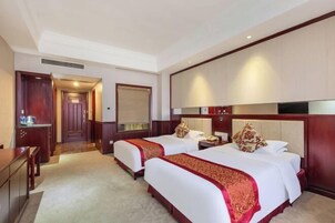 Quarto de hotel com duas camas, uma escrivaninha, uma cadeira, um telefone e uma janela com persianas.