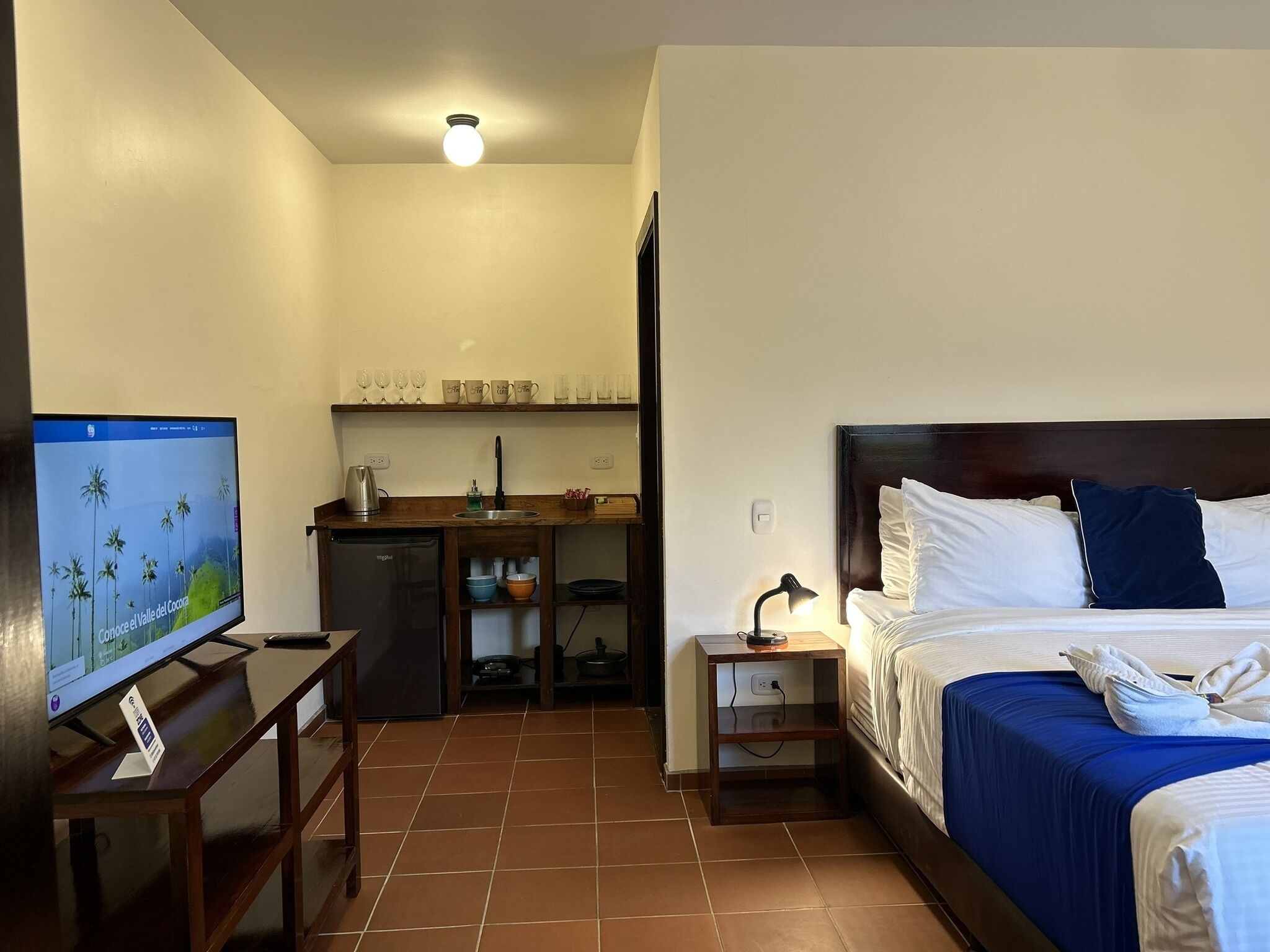 Quarto de hotel pequeno com cama, mesa de cabeceira, escrivaninha com computador, pequena cozinha e televisão na escrivaninha.