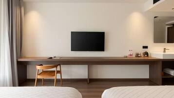 Quarto de hotel com cama, escrivaninha com cadeira, televisão fixada na parede e espelho sobre a pia.