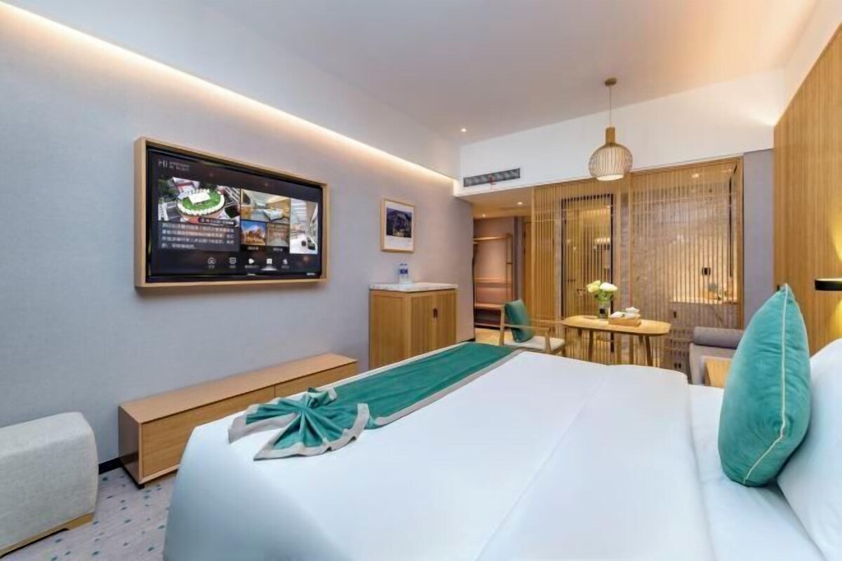 Quarto de hotel moderno com uma cama grande, uma mesa de cabeceira, uma pequena escrivaninha, uma cadeira, uma TV fixada na parede e um painel de madeira.