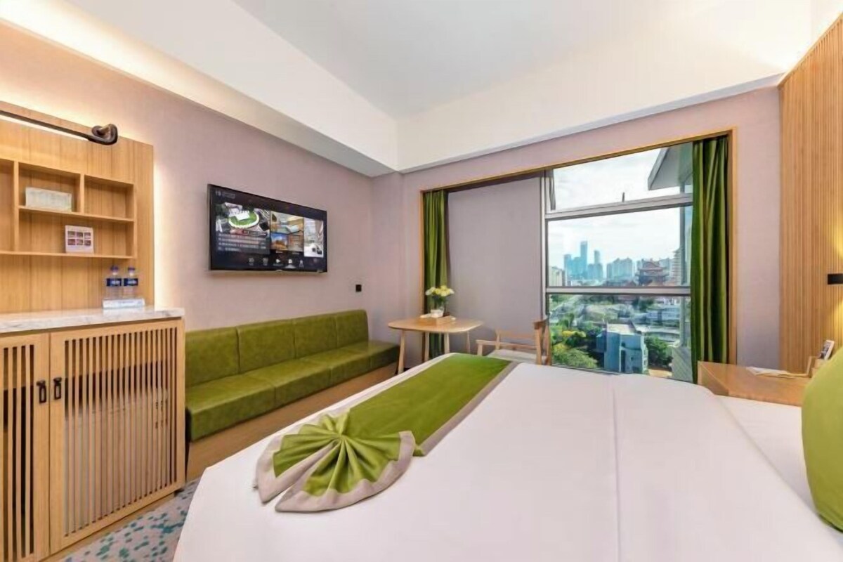 Quarto de hotel com uma cama, um sofá verde, uma TV, uma mesinha com um vaso e uma janela com vista para a cidade.