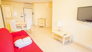 Quarto de hotel com sofá vermelho, uma mesinha com abajur, televisão na parede, escrivaninha com cadeiras e um armário.