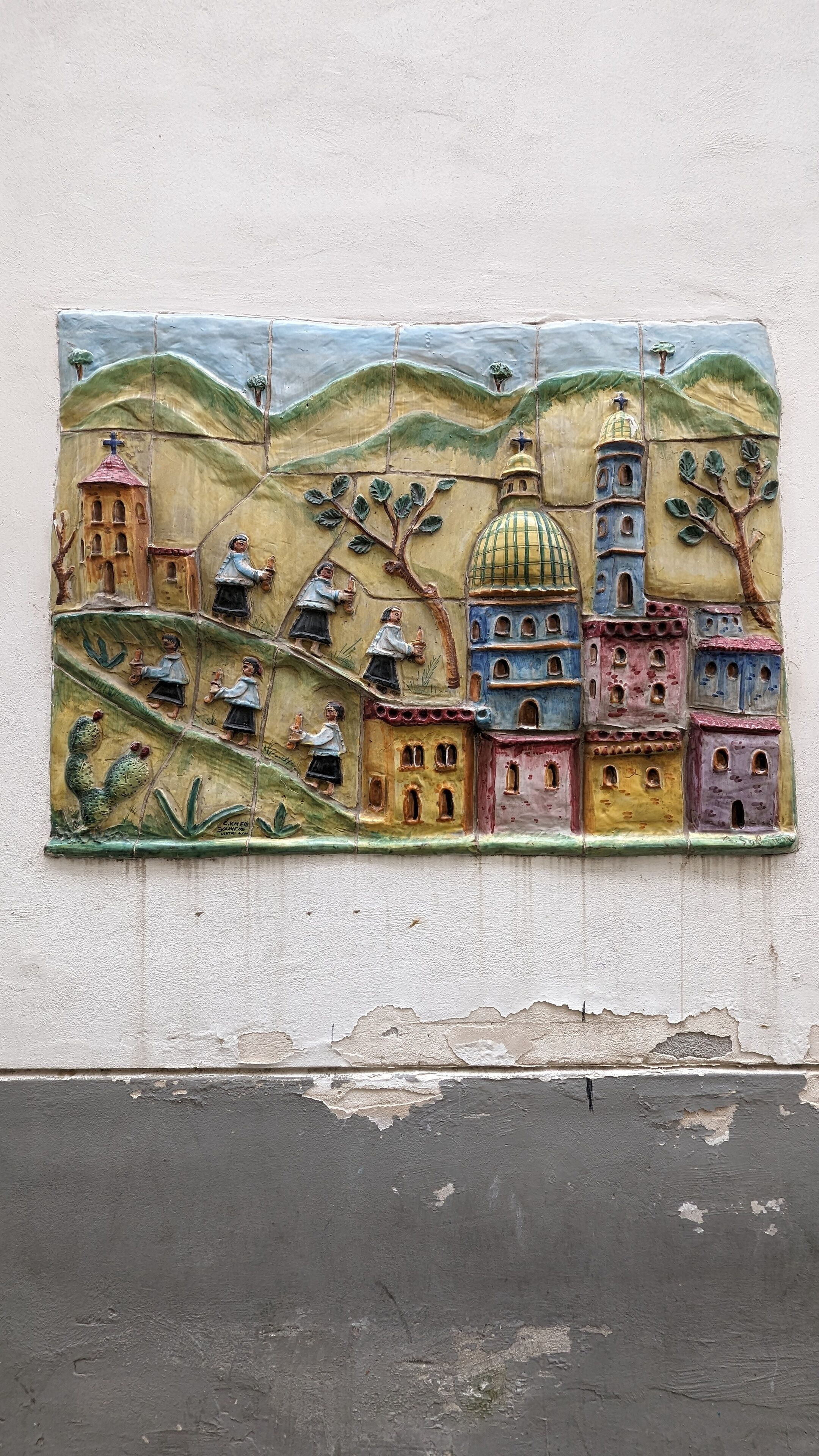 Um mural de mosaico colorido numa parede retrata uma cena de vila com edifícios, árvores e uma igreja.