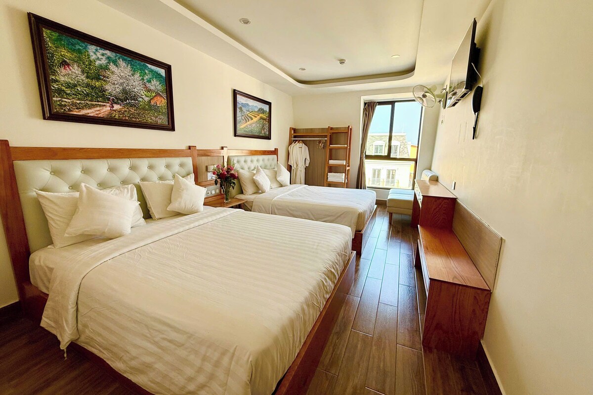Quarto de hotel com duas camas, cabeceira de madeira, uma janela grande e um quadro na parede.