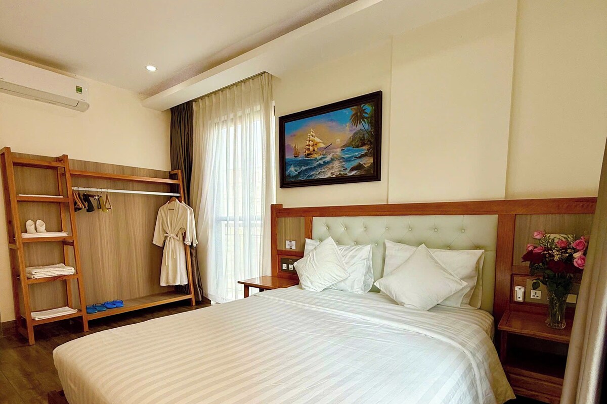 Quarto de hotel com uma cama grande, um guarda-roupa de madeira, um quadro na parede e uma mesa de cabeceira com um vaso.