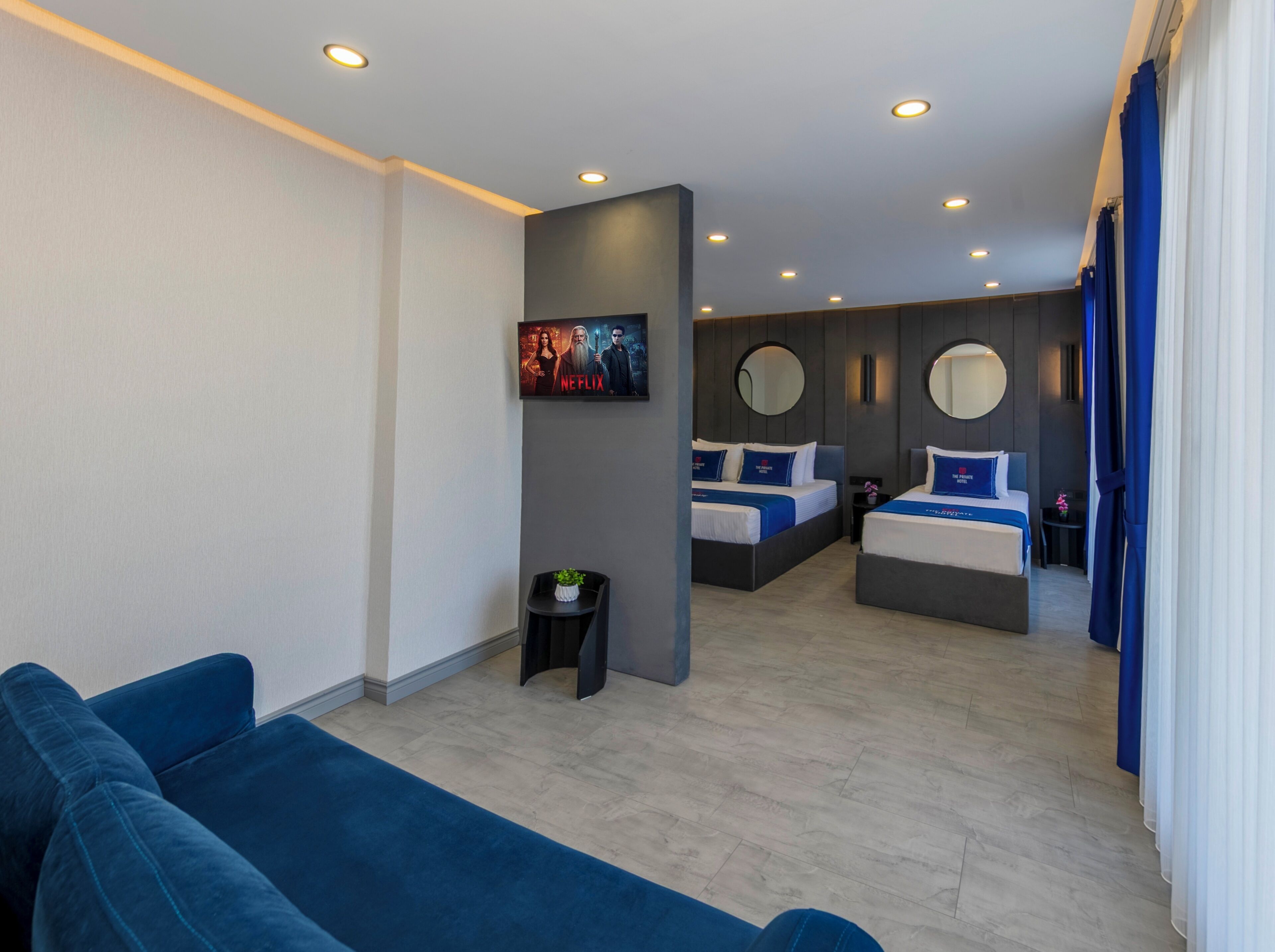 Quarto de hotel moderno com duas camas, um sofá, uma mesa pequena e uma televisão fixada na parede.