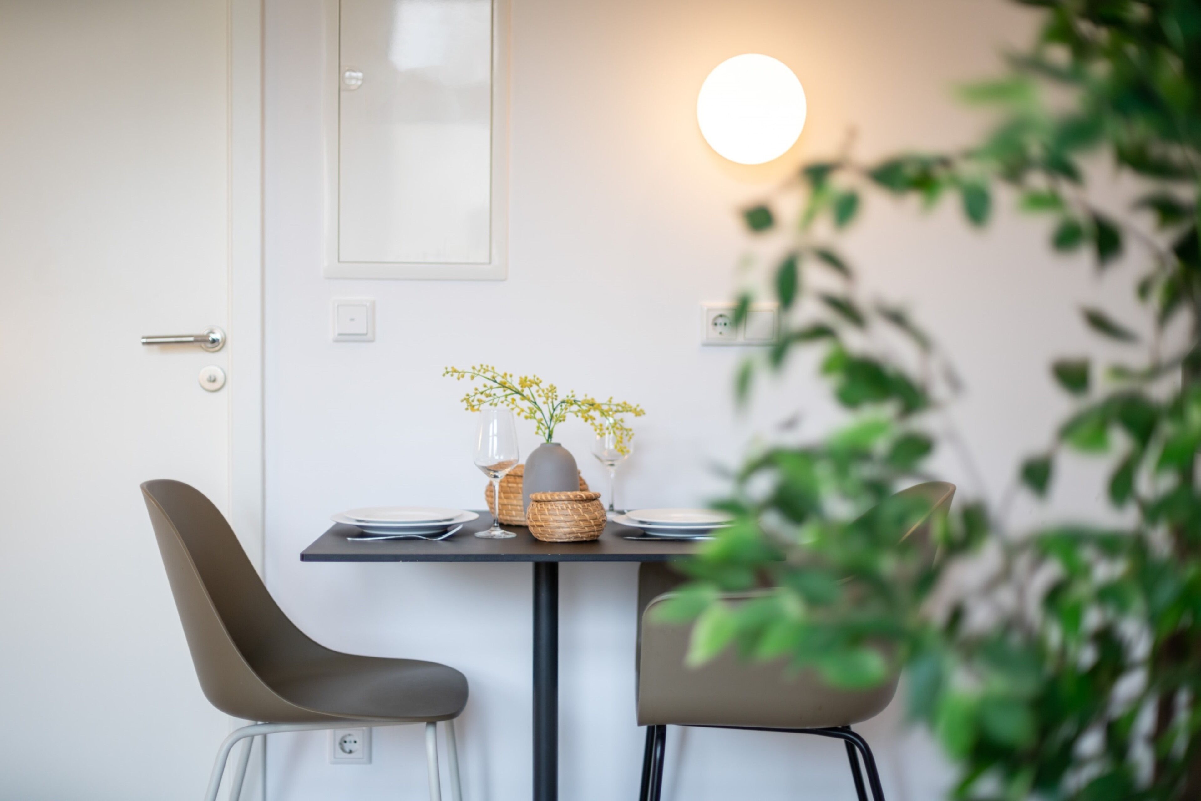 Uma sala de jantar com duas cadeiras, uma mesa, um vaso com flores e uma luminária na parede.
