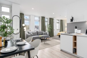 Uma sala de estar moderna com um sofá cinzento, mesa de jantar posta com pratos e copos, e uma cozinha com bancada e estantes.