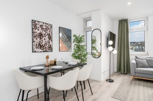 Uma sala de jantar moderna com uma mesa preta, cadeiras brancas e uma planta em vaso. Há um sofá e uma janela com cortinas na área de estar adjacente.