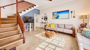 Uma sala de estar ampla com escada de madeira, ventilador de teto, uma pintura grande e um sofá com almofadas.