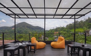 Um terraço com mesas e cadeiras de madeira, um bean bag amarelo e vista para uma paisagem montanhosa.