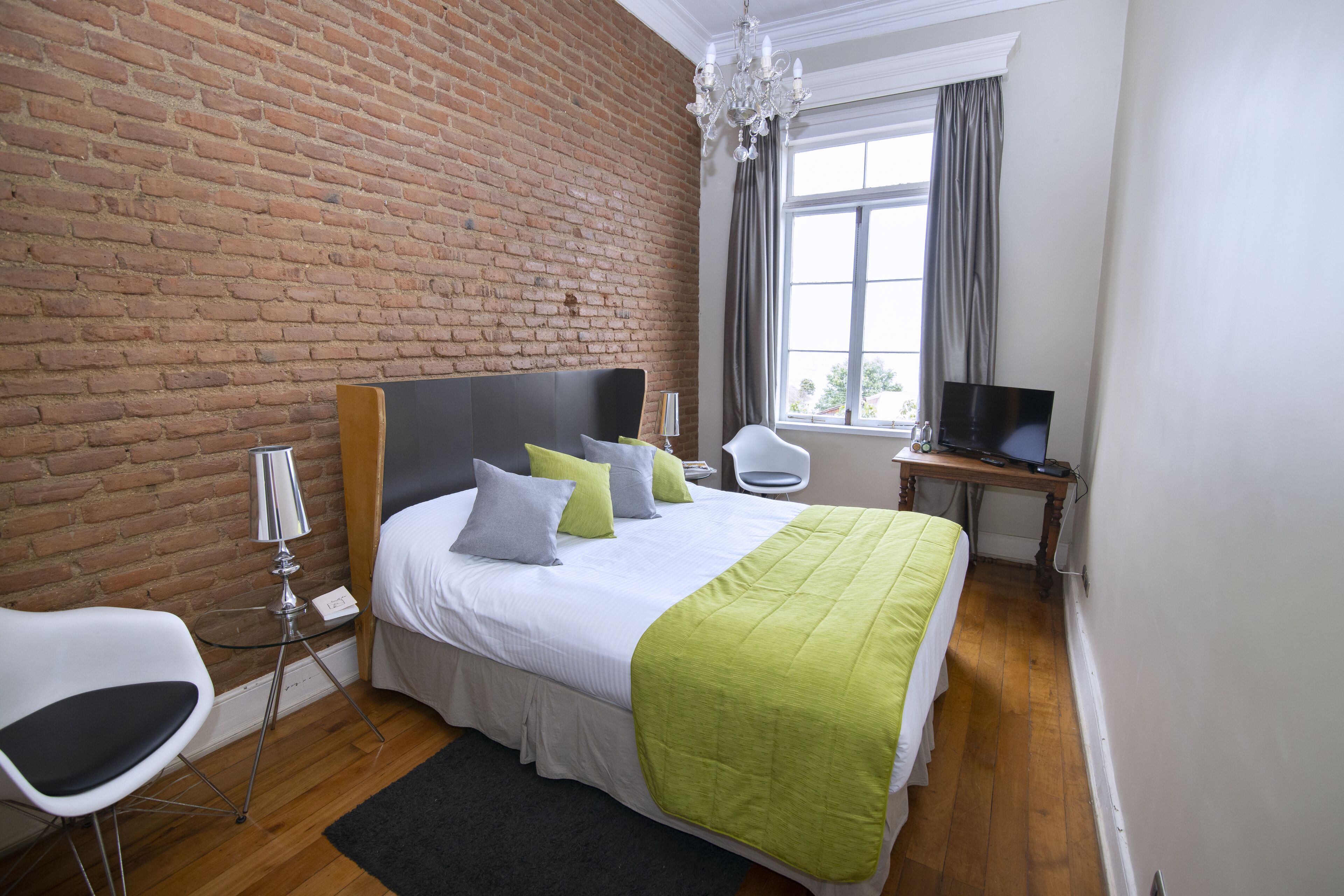 Quarto com parede de tijolos, cama com colcha verde e branca, cadeira branca, escrivaninha com computador e janela com cortinas.
