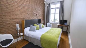 Quarto com parede de tijolos, cama com colcha verde e branca, cadeira branca, escrivaninha com computador e janela com cortinas.