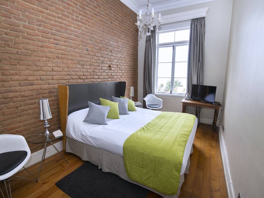 Quarto com parede de tijolos, cama com colcha verde e branca, cadeira branca, escrivaninha com computador e janela com cortinas.