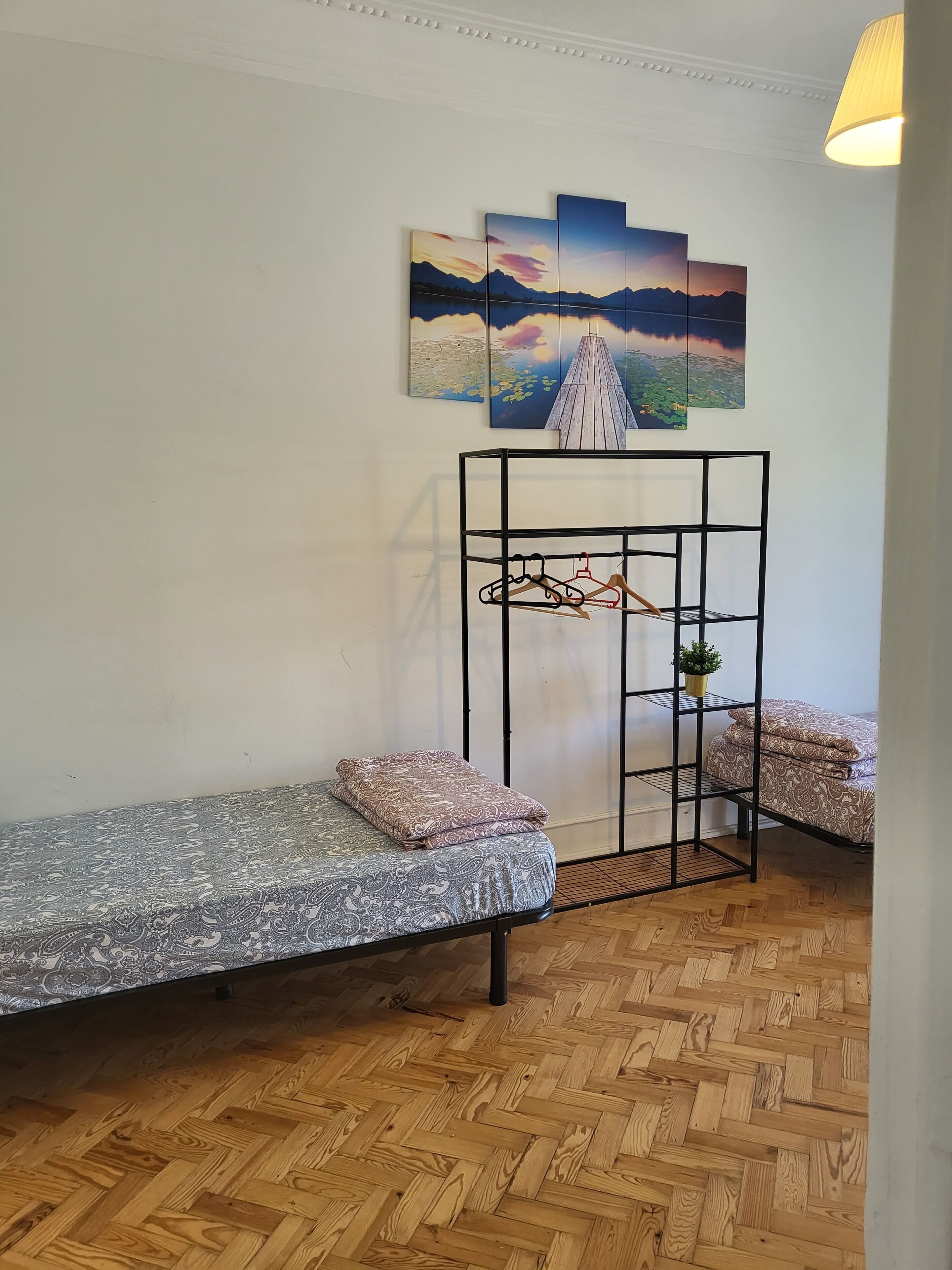 Quarto com piso de madeira em padrão herringbone, cama com roupa de cama estampada, estante de metal preta com uma planta, e três quadros na parede.
