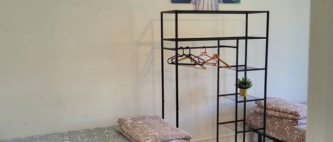 Quarto com piso de madeira em padrão herringbone, cama com roupa de cama estampada, estante de metal preta com uma planta, e três quadros na parede.