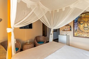 Quarto com cama de dossel, cadeiras de vime, uma mesa pequena e um quadro na parede.