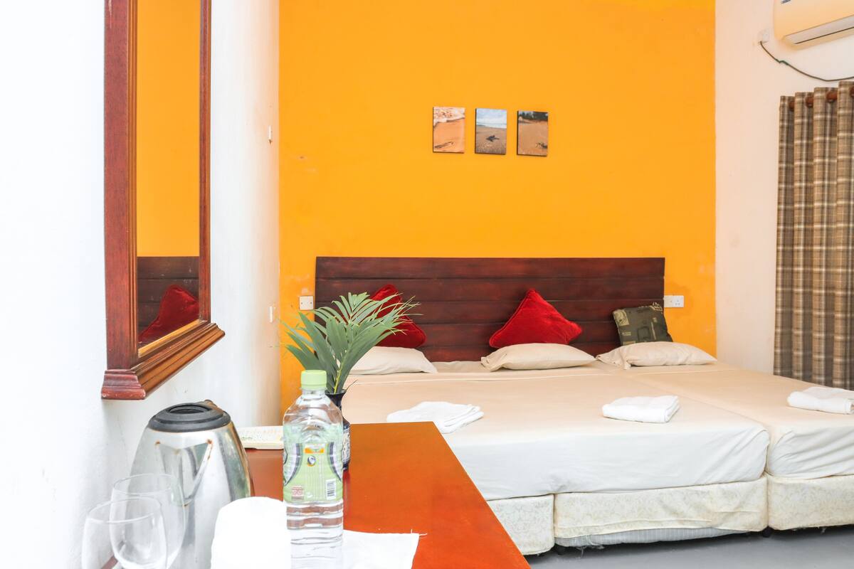 Quarto de hotel com cama, cabeceira de madeira, espelho, mesa com garrafa e copos, e parede com três quadros.