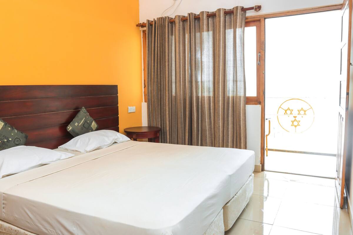 Quarto de hotel com cama, mesa redonda pequena, cabeceira de madeira e porta de vidro deslizante que dá para uma varanda.