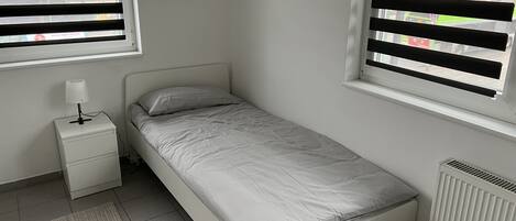Quarto pequeno com uma cama de solteiro, uma mesa de cabeceira, uma lâmpada e um radiador. Há duas janelas com persianas, e um quadro na parede.