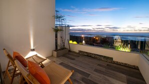 Uma varanda com vista para o oceano ao entardecer, com uma cadeira de madeira, uma mesa pequena e um vaso de planta.