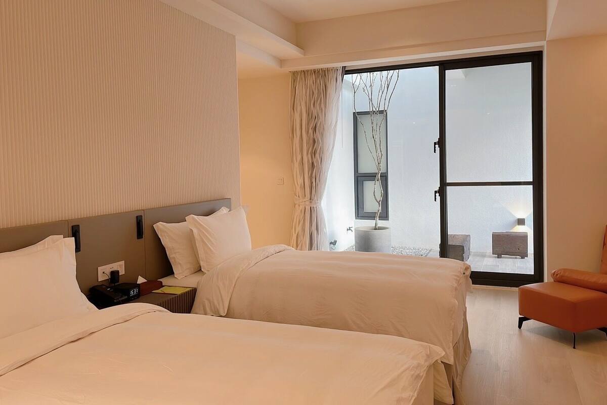 Quarto de hotel com duas camas, uma cadeira, uma mesinha de cabeceira e vista para o exterior através de uma porta de vidro.