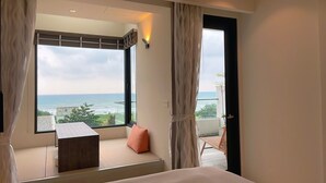 Quarto com uma grande janela com vista para o mar, uma cama, uma mesinha e um banco acolchoado.