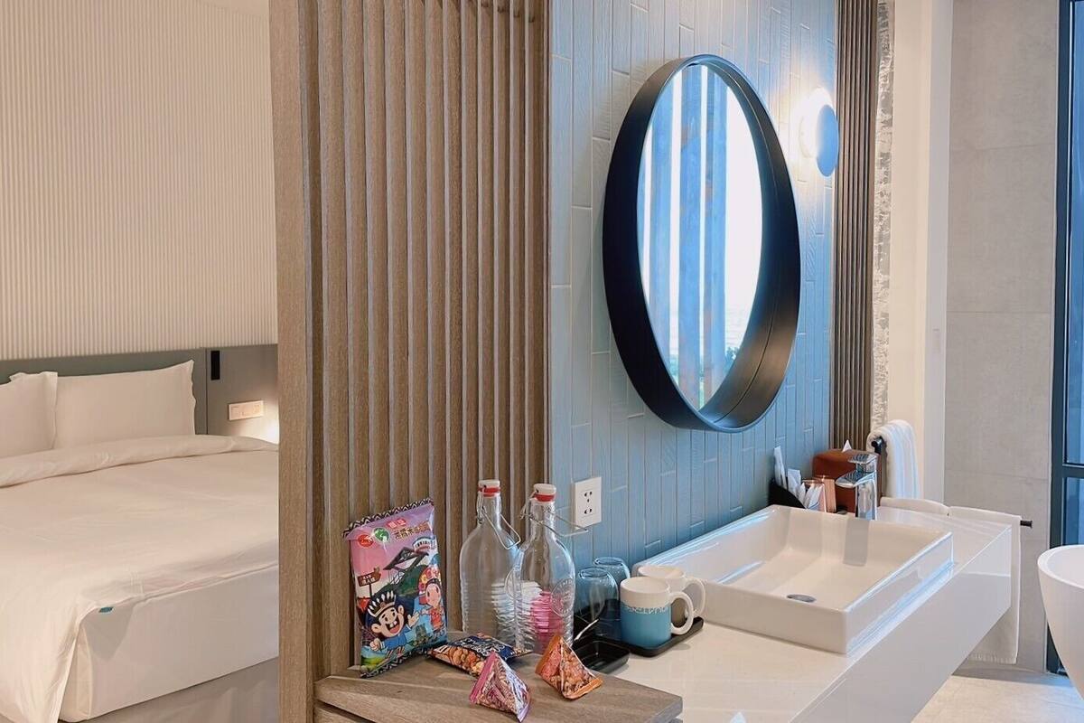 Quarto de hotel com cama, mesa de cabeceira com produtos de higiene pessoal, espelho e janela com persianas.