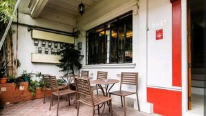 Uma área externa com mesas e cadeiras de madeira, uma planta em vaso e entrada de prédio com porta vermelha e número 2932.