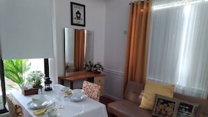 Uma área de refeições com uma mesa posta para quatro pessoas, um sofá com almofadas decorativas, um espelho e uma janela com cortinas.