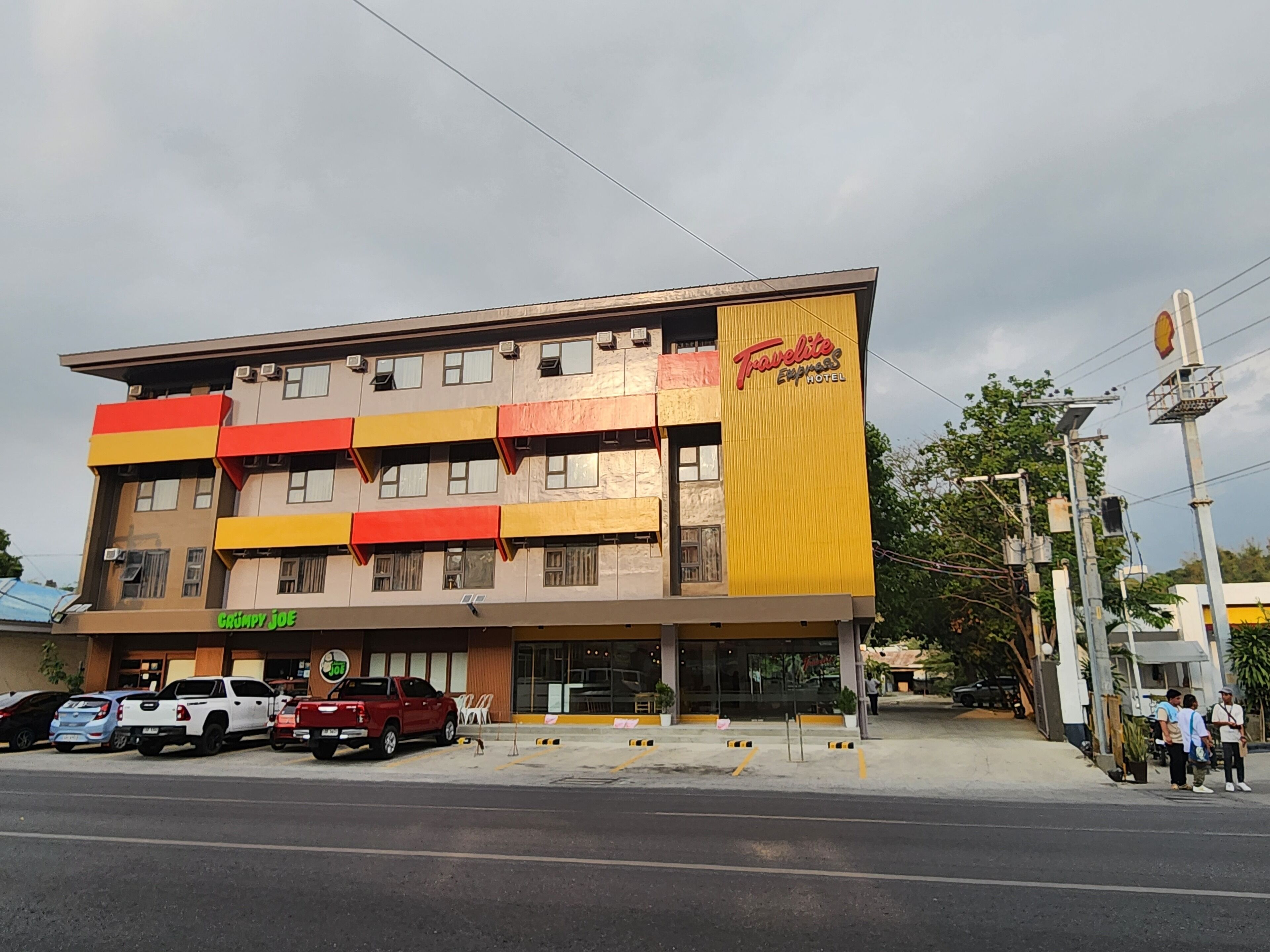 Um edifício de vários andares com fachada em amarelo e vermelho marcante, exibindo a placa 'Travellite Express Hotel'. Há vários veículos estacionados na frente, incluindo uma caminhonete vermelha e um SUV branco.