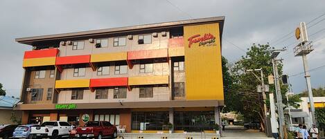 Um edifício de vários andares com fachada em amarelo e vermelho marcante, exibindo a placa 'Travellite Express Hotel'. Há vários veículos estacionados na frente, incluindo uma caminhonete vermelha e um SUV branco.