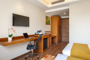 Quarto de hotel com escrivaninha de madeira, um laptop, uma cadeira, uma cama com manta amarela, uma televisão e um quadro na parede.