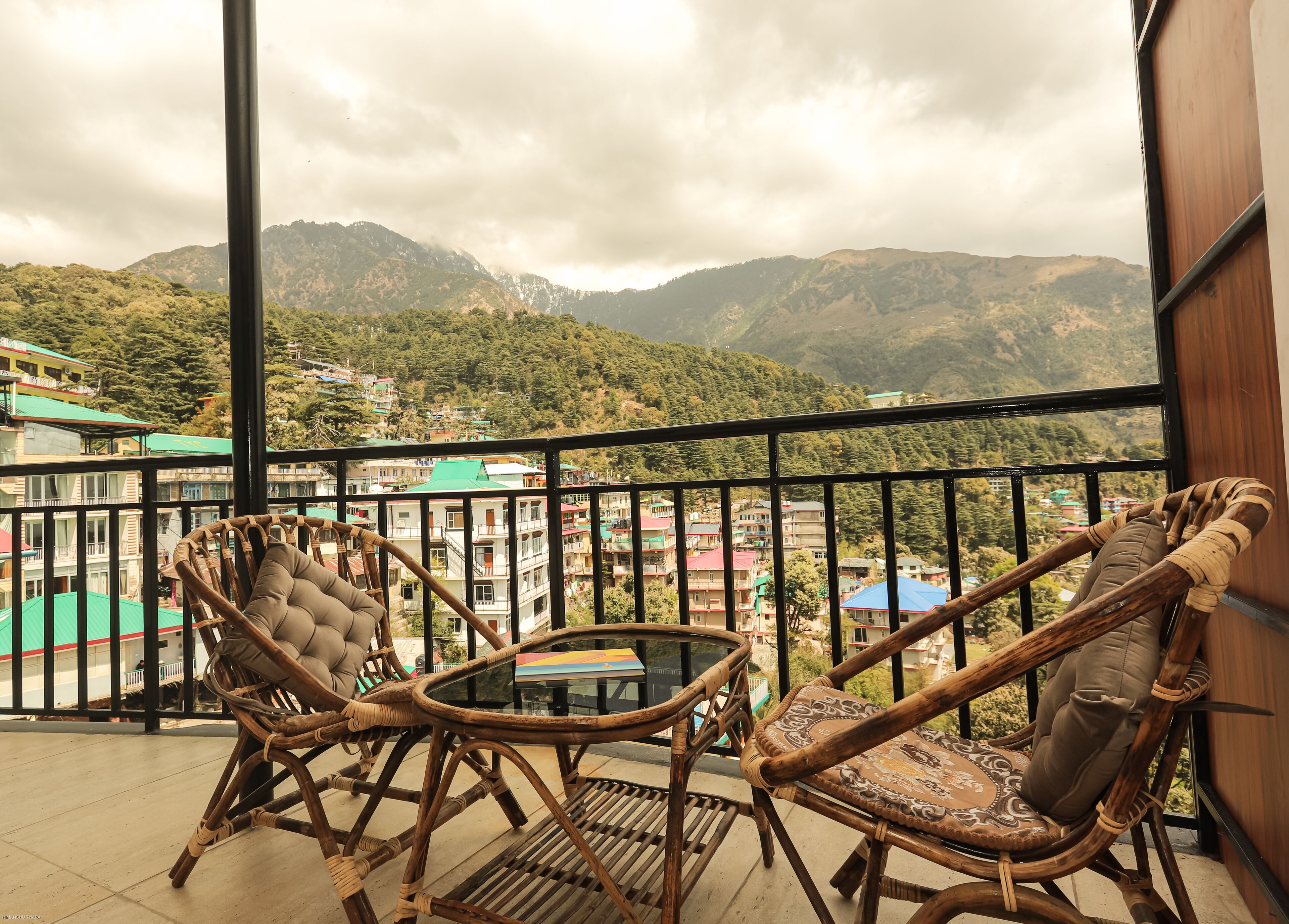 Uma varanda com cadeiras de vime e uma mesa pequena, com vista para uma paisagem montanhosa e edifícios ao fundo.