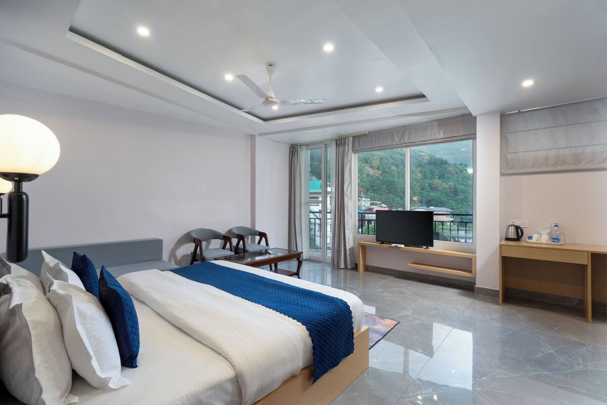 Quarto de hotel moderno com uma cama grande, uma escrivaninha, uma televisão e uma varanda com vista para o verde.
