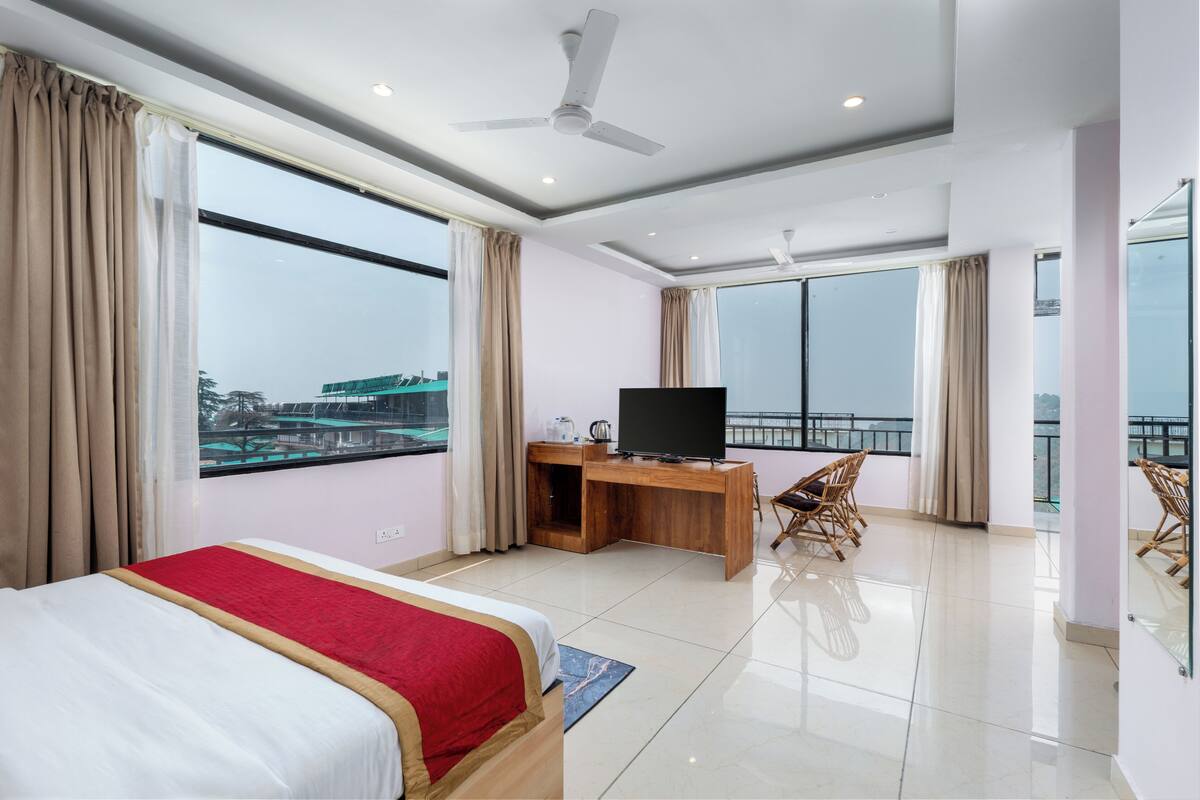 Quarto de hotel moderno com uma cama grande, uma mesa de trabalho com uma TV e uma varanda com vista para uma ponte.