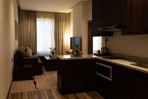 Uma sala de estar moderna com um sofá, uma mesa de centro e uma televisão. Há uma área de cozinha com micro-ondas e uma cesta de frutas.