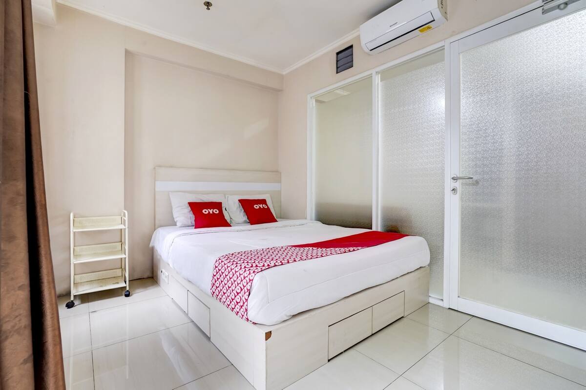 Cama bem arrumada com manta branca e vermelha, mesa de cabeceira branca com travesseiro vermelho e ar-condicionado branco fixado na parede.