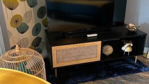 Uma sala de estar moderna com uma TV de tela plana sobre um suporte preto, uma parede decorativa com padrões de folhas, uma mesinha lateral trançada e um tapete felpudo azul.