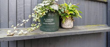 Uma prateleira de madeira com duas plantas em vasos, uma delas com a etiqueta "FLOWERS MARKET FRESH PRODUCT" e outra com vaso decorativo.