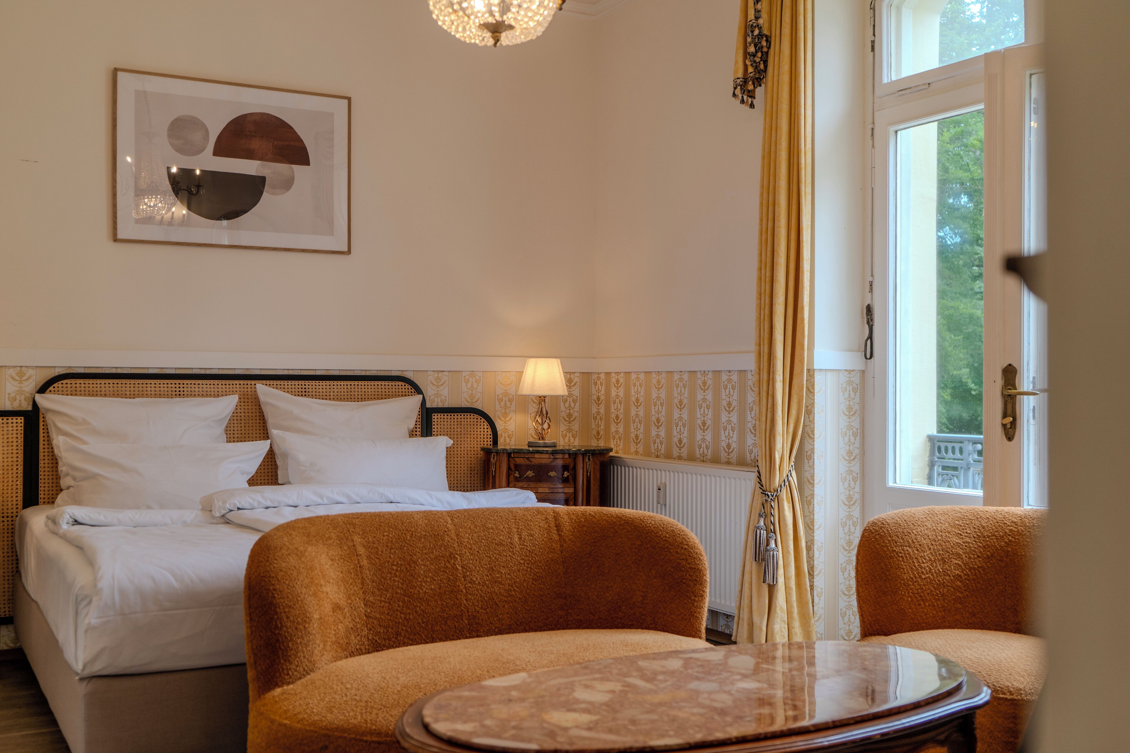 Quarto de hotel com uma cama, duas cadeiras, uma mesa, um abajur, um quadro na parede e uma janela com cortinas.