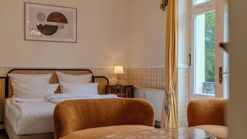 Quarto de hotel com uma cama, duas cadeiras, uma mesa, um abajur, um quadro na parede e uma janela com cortinas.