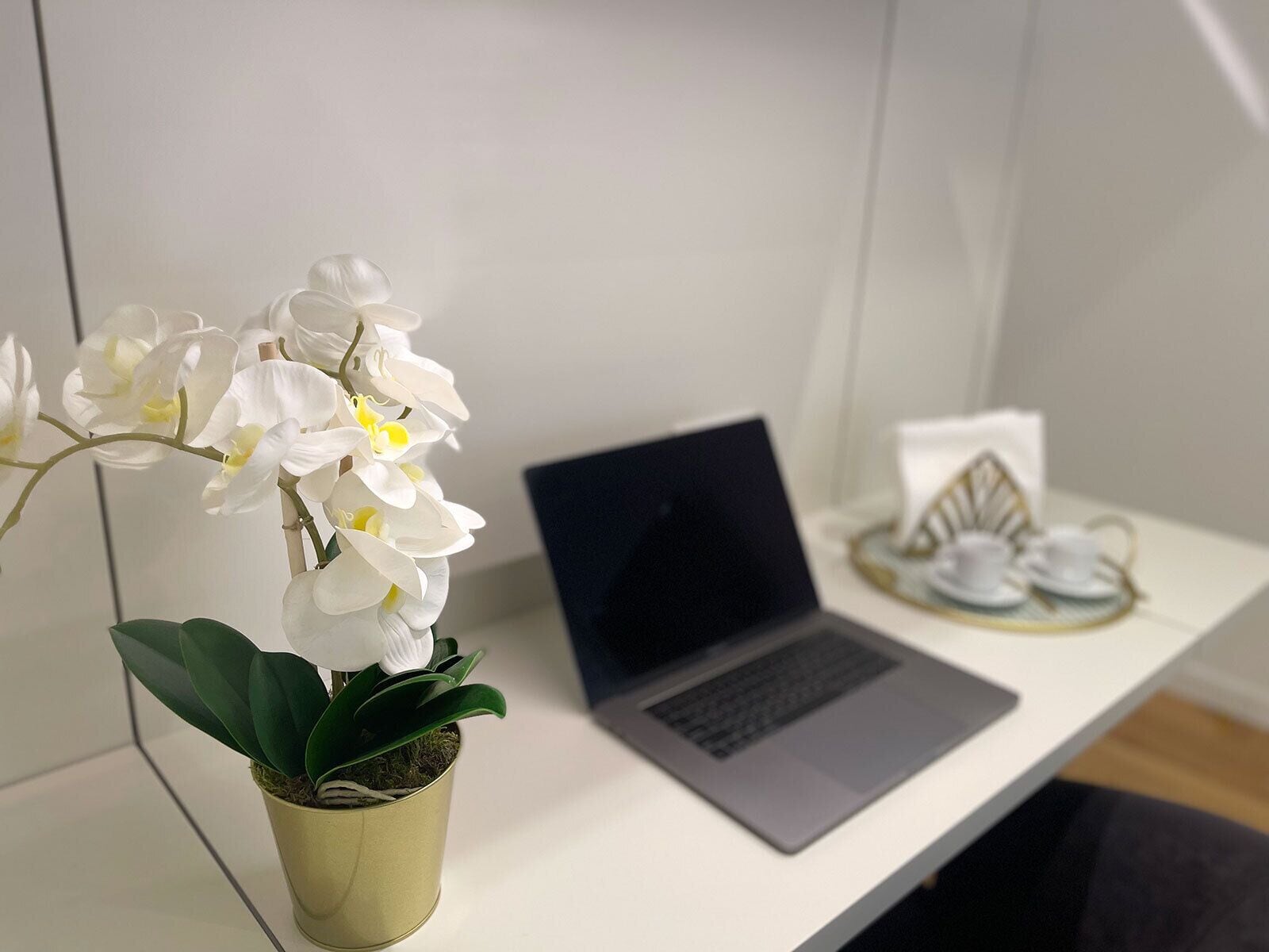 Uma mesa com um laptop, uma orquídea em vaso e um objeto decorativo com xícaras e pireses.