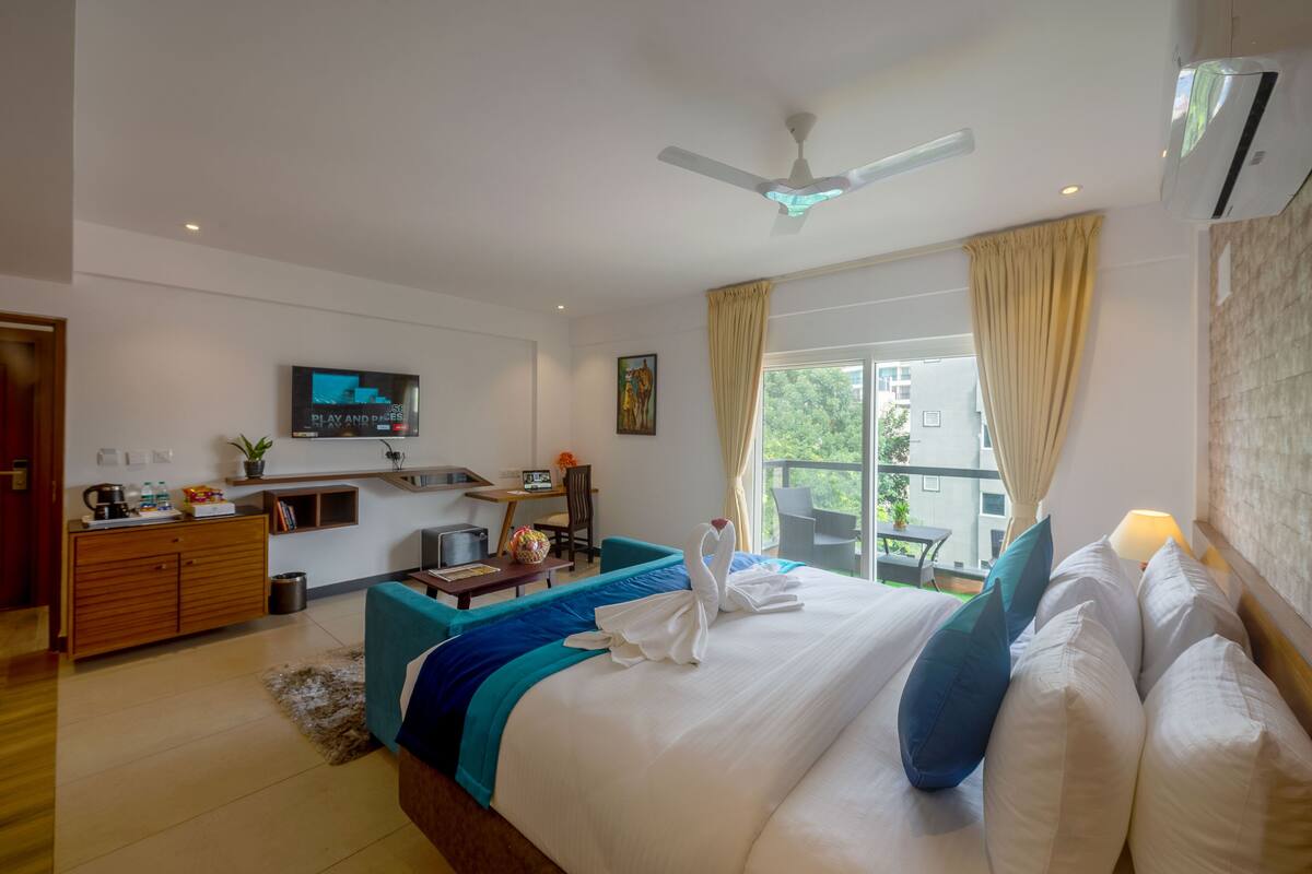 Quarto de hotel moderno com uma cama grande, uma escrivaninha com computador, televisão e varanda com vista para a cidade.