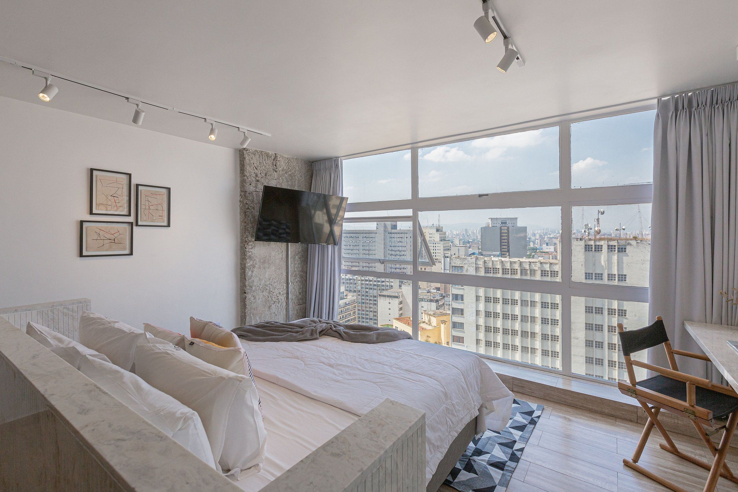 Quarto moderno com uma cama grande, televisão na parede e vista para a cidade através de amplas janelas.