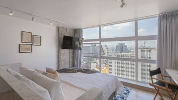 Quarto moderno com uma cama grande, televisão na parede e vista para a cidade através de amplas janelas.