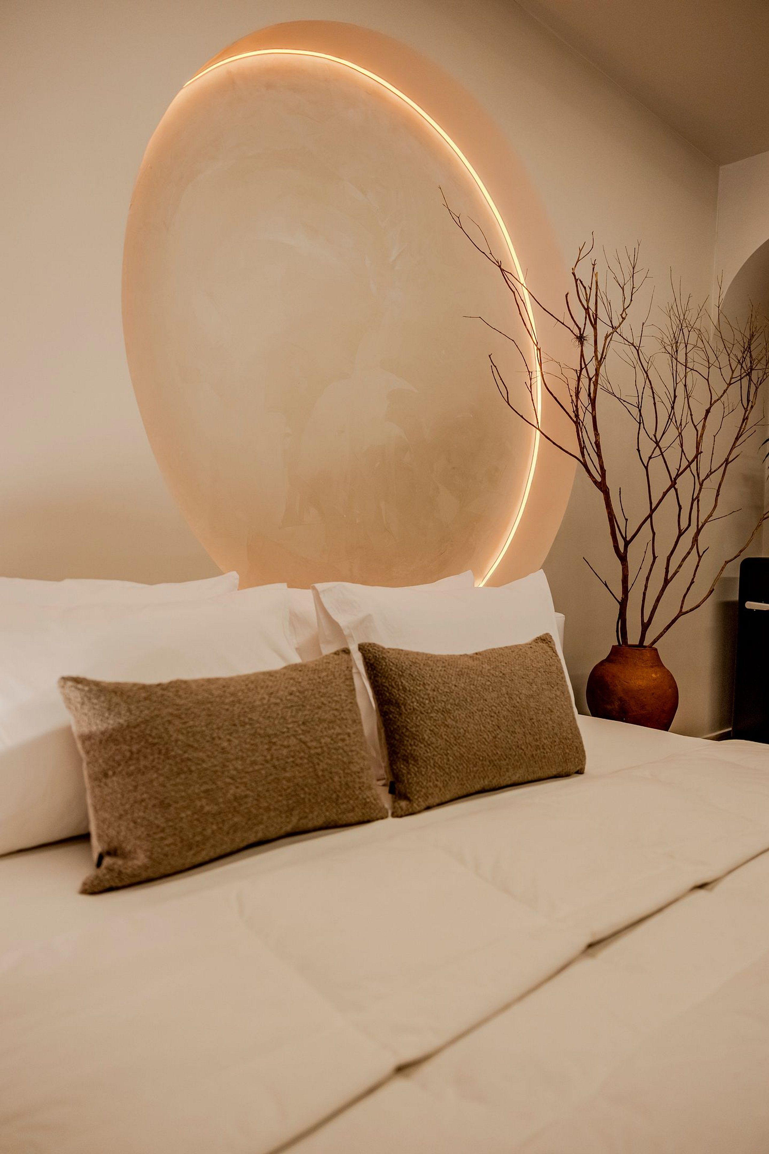 Cama com roupa branca, duas almofadas castanhas, um vaso com ramos e um candeeiro fixado na parede.